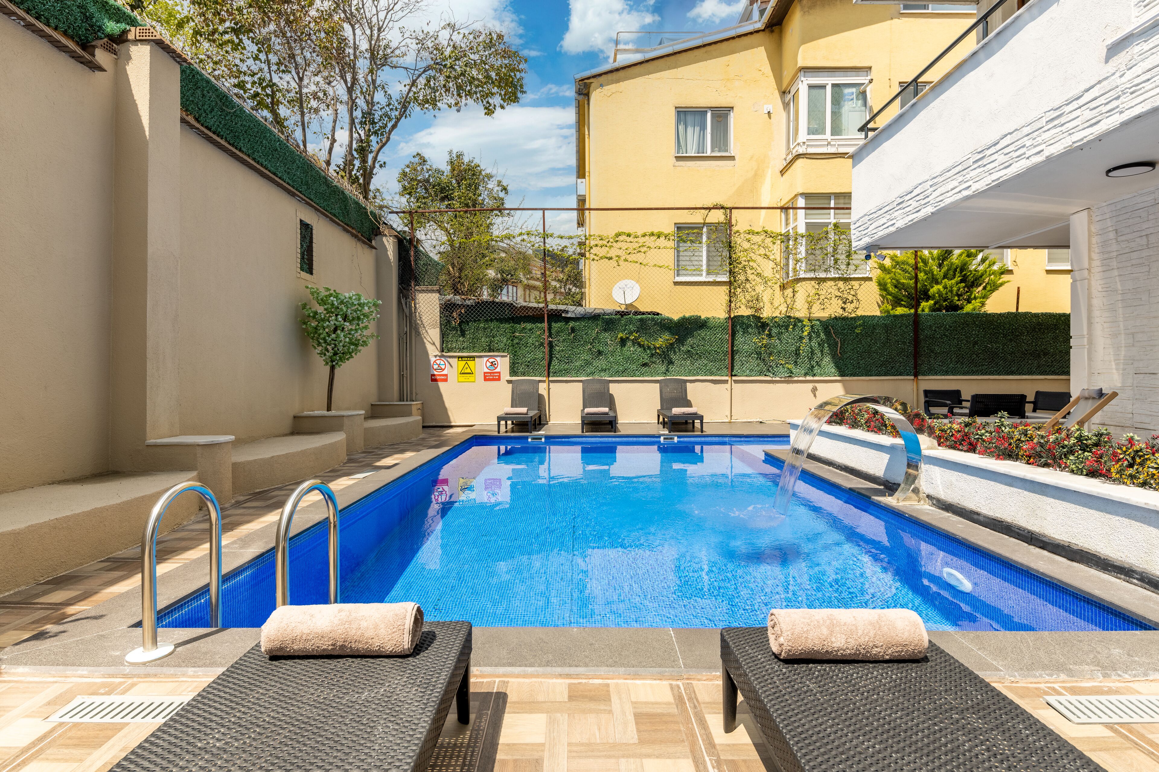 Uma piscina com duas espreguiçadeiras, um elemento aquático e um edifício com um muro verde ao fundo.