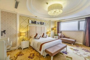 Quarto de hotel com cama grande, um banco, mesa de cabeceira, luminária e uma janela com cortinas.