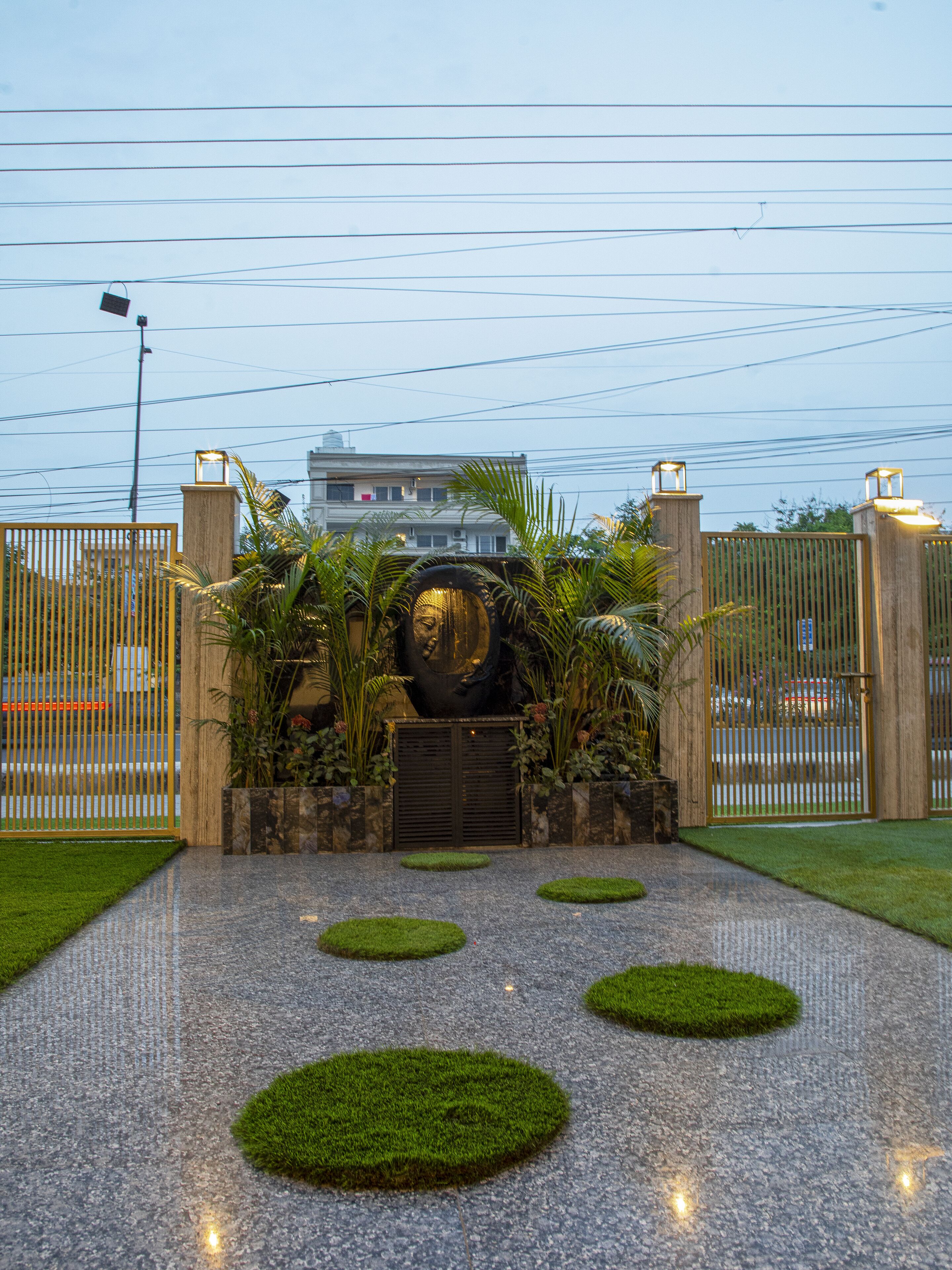 Uma entrada com portão e uma escultura de metal decorativa, um caminho pavimentado com áreas de gramado circulares, e um edifício ao fundo.