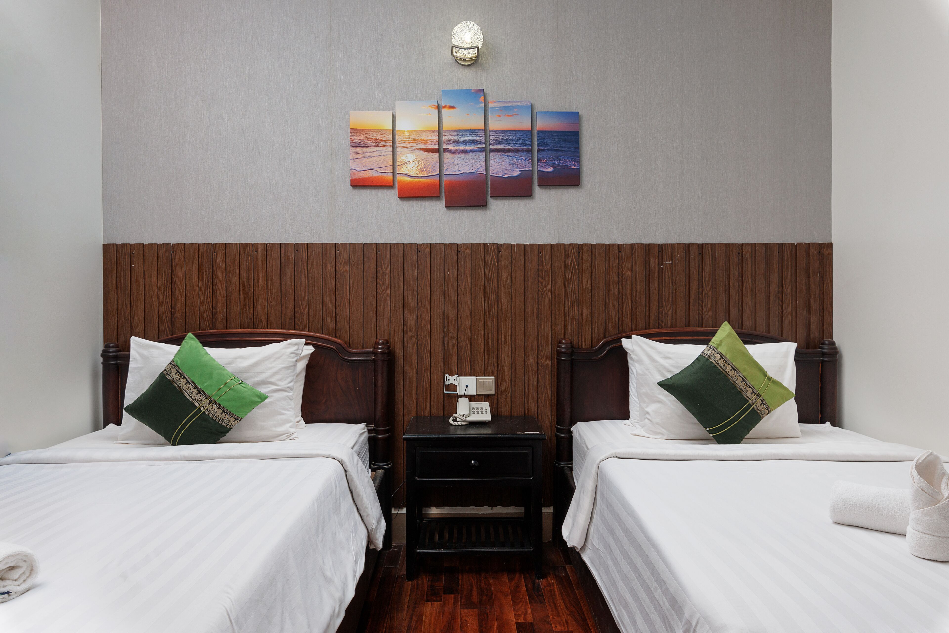 Quarto de hotel com duas camas, uma mesa de cabeceira com telefone e uma obra de arte na parede retratando um pôr do sol sobre o oceano.