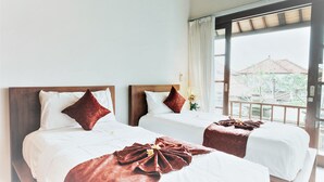 Quarto de hotel com duas camas, roupa de cama branca, almofadas decorativas castanhas e uma varanda com vista para um edifício e vegetação.