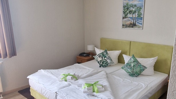Uma cama bem arrumada com lençóis brancos e detalhes verdes, um quadro de uma árvore na parede e piso de madeira.