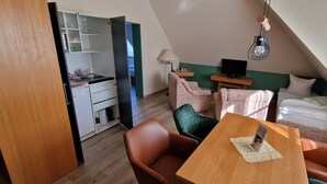 Quarto com mesa de jantar, duas cadeiras, cama com almofadas, televisão em um suporte e cozinha visível por uma porta aberta.
