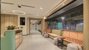 Uma área de recepção moderna com balcão verde claro, assentos com almofadas estampadas e uma grande janela com vista para o exterior.