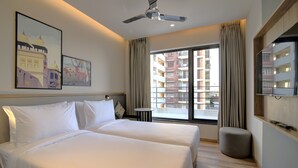 Quarto de hotel com duas camas, um ventilador de teto, uma janela grande com cortinas e obras de arte na parede.