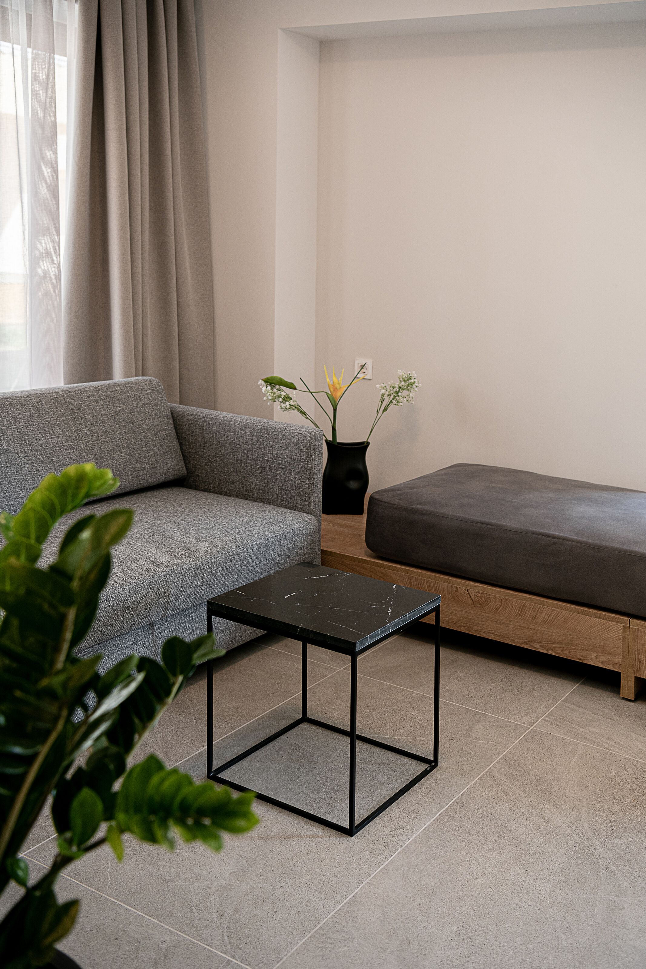 Uma sala de estar moderna com um sofá cinzento, um banco de madeira, uma mesa de centro preta e um vaso com flores.