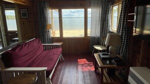 Interior de cabana com teto de madeira, uma cama, uma mesa com abajur, televisão e vista para a praia.