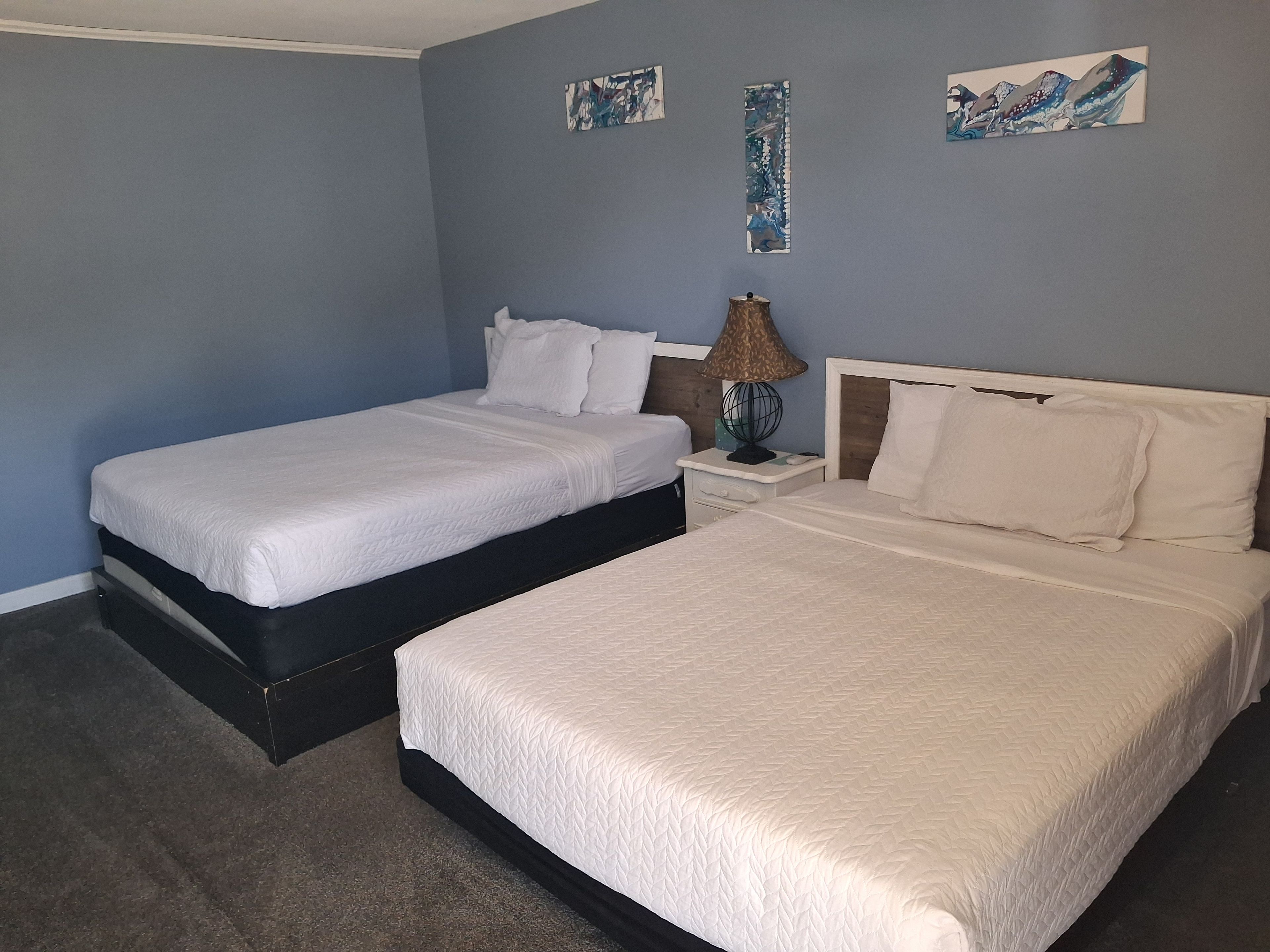 Quarto de hotel com duas camas, mesa de cabeceira com abajur e obras de arte nas paredes.
