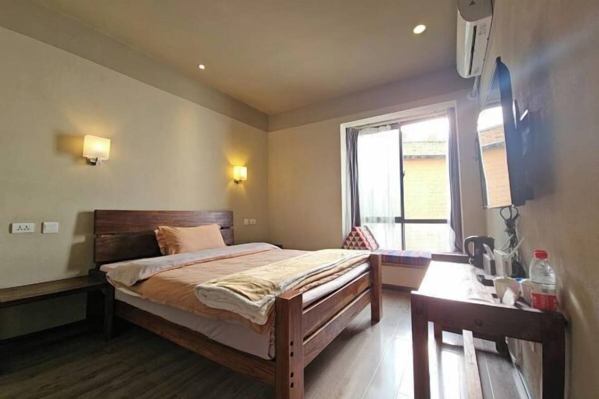 Um quarto com cama de madeira, duas luminárias fixadas na parede, uma janela com cortinas, uma escrivaninha com cadeira e um ar-condicionado fixado na parede.