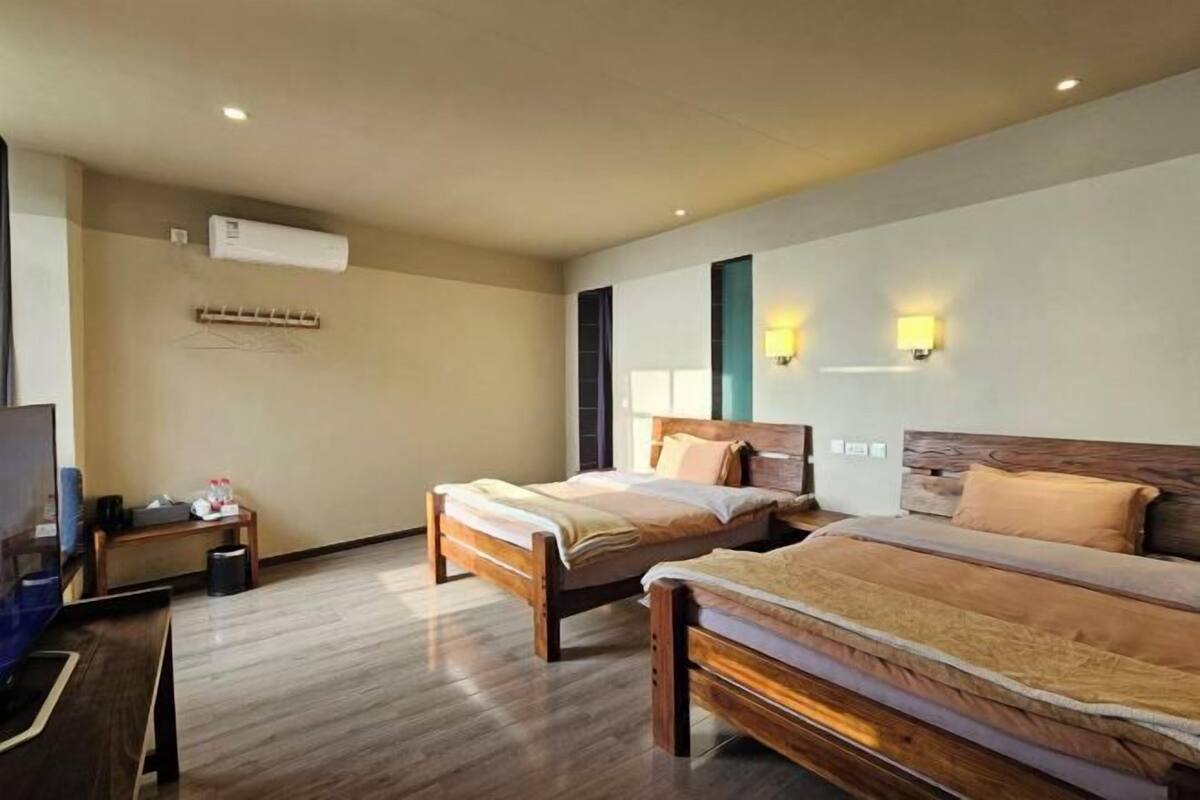 Quarto de hotel com duas camas de madeira, uma escrivaninha com cadeira, ar condicionado e uma televisão.