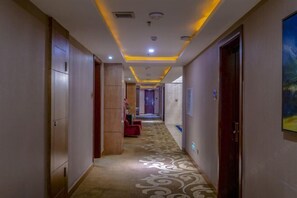 Corredor de hotel com carpete estampado, iluminação embutida e uma porta à direita.