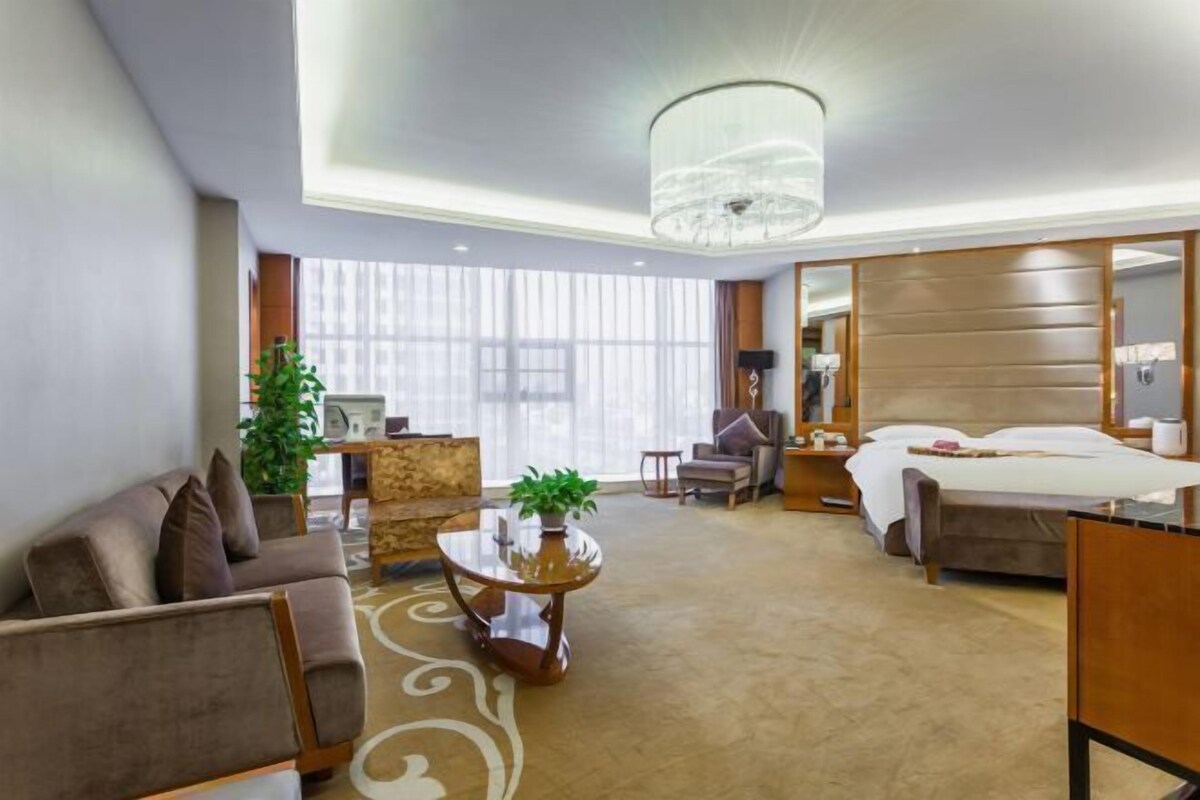 Quarto de hotel com cama, sofá, escrivaninha com cadeira, mesa pequena com planta e uma janela grande com cortinas.