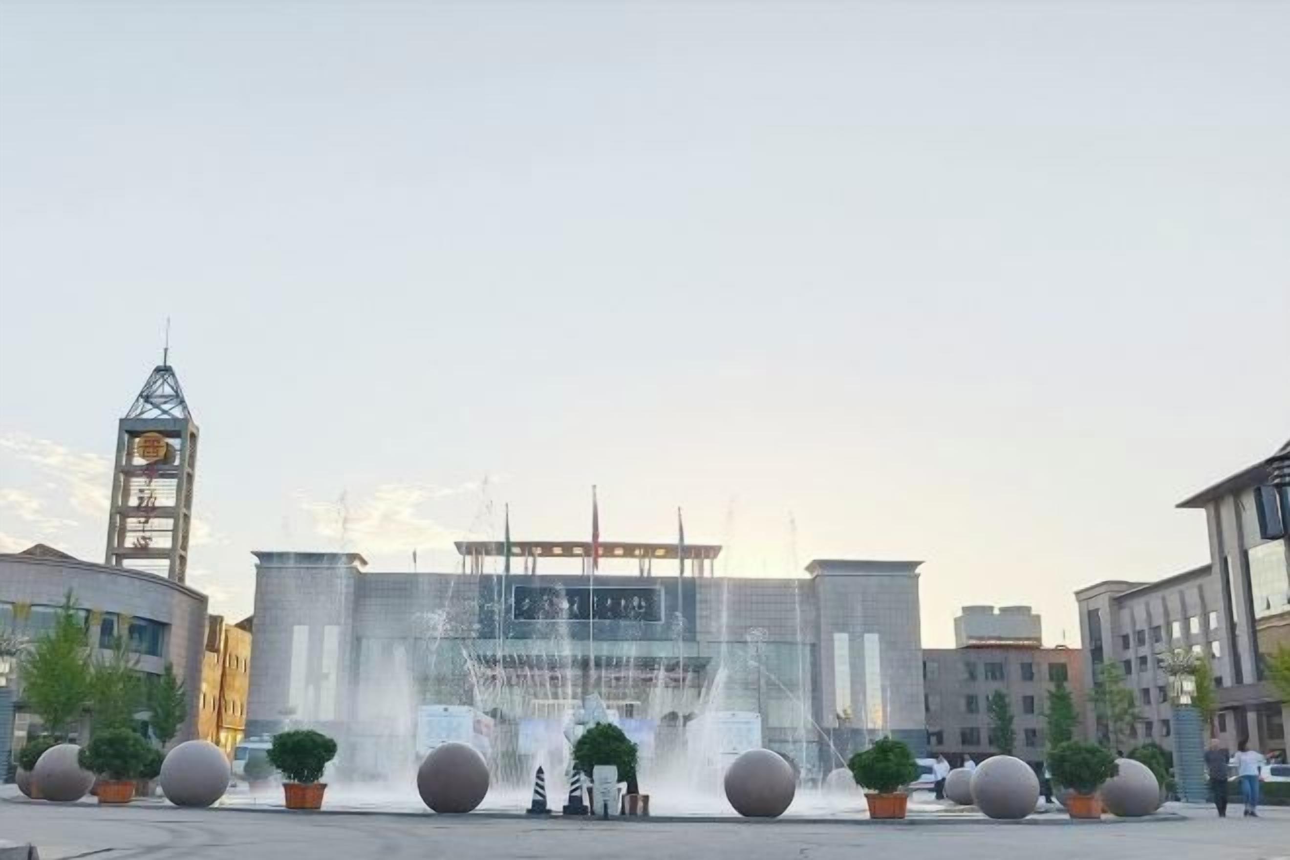 Uma praça pública com uma fonte, plantas em vasos e um campanário.
