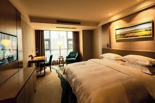 Quarto de hotel com uma cama grande, uma mesa, uma cadeira, uma televisão e um quadro na parede.