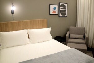 Quarto de hotel com cama, uma cadeira, uma luminária na parede e duas obras de arte emolduradas.