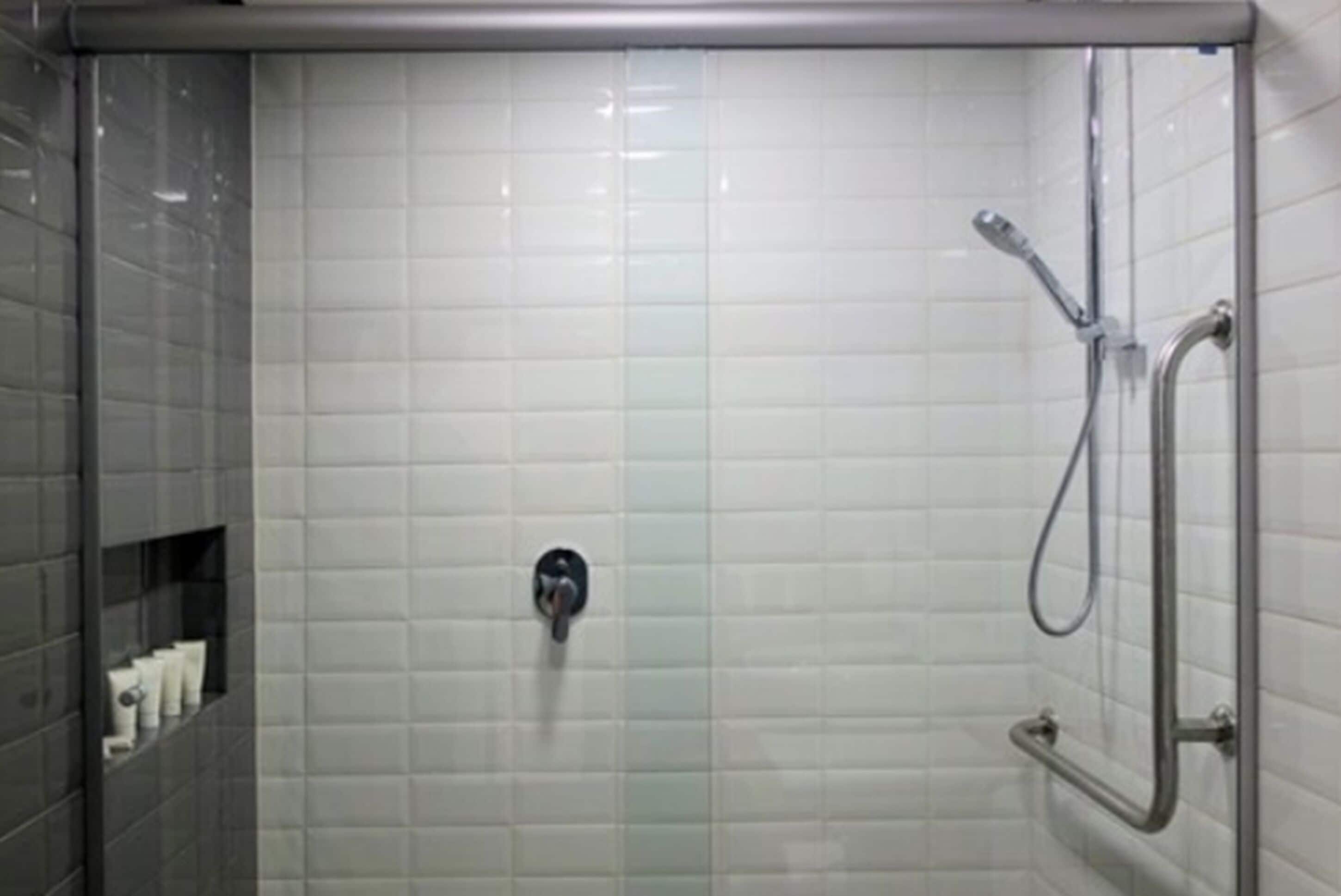Área de chuveiro com paredes azulejadas em branco, porta de vidro e chuveiro de metal.