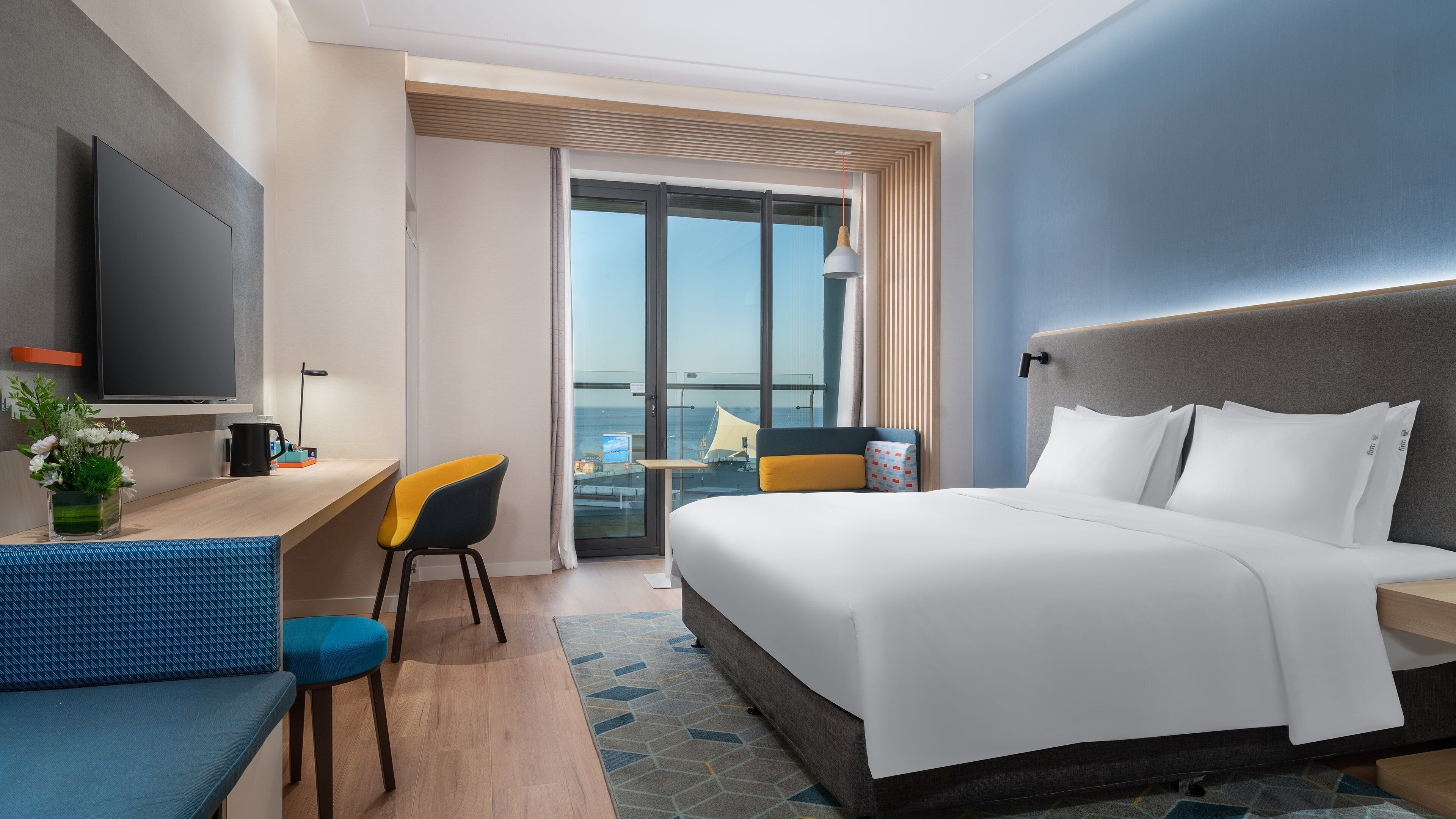 Quarto de hotel moderno com cama grande, escrivaninha, cadeira, televisão e varanda com vista para o mar.