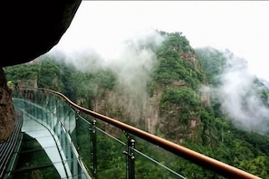 Uma ponte de vidro com grades de metal e corrimão de madeira, com vista para uma montanha verde e exuberante envolta em névoa.