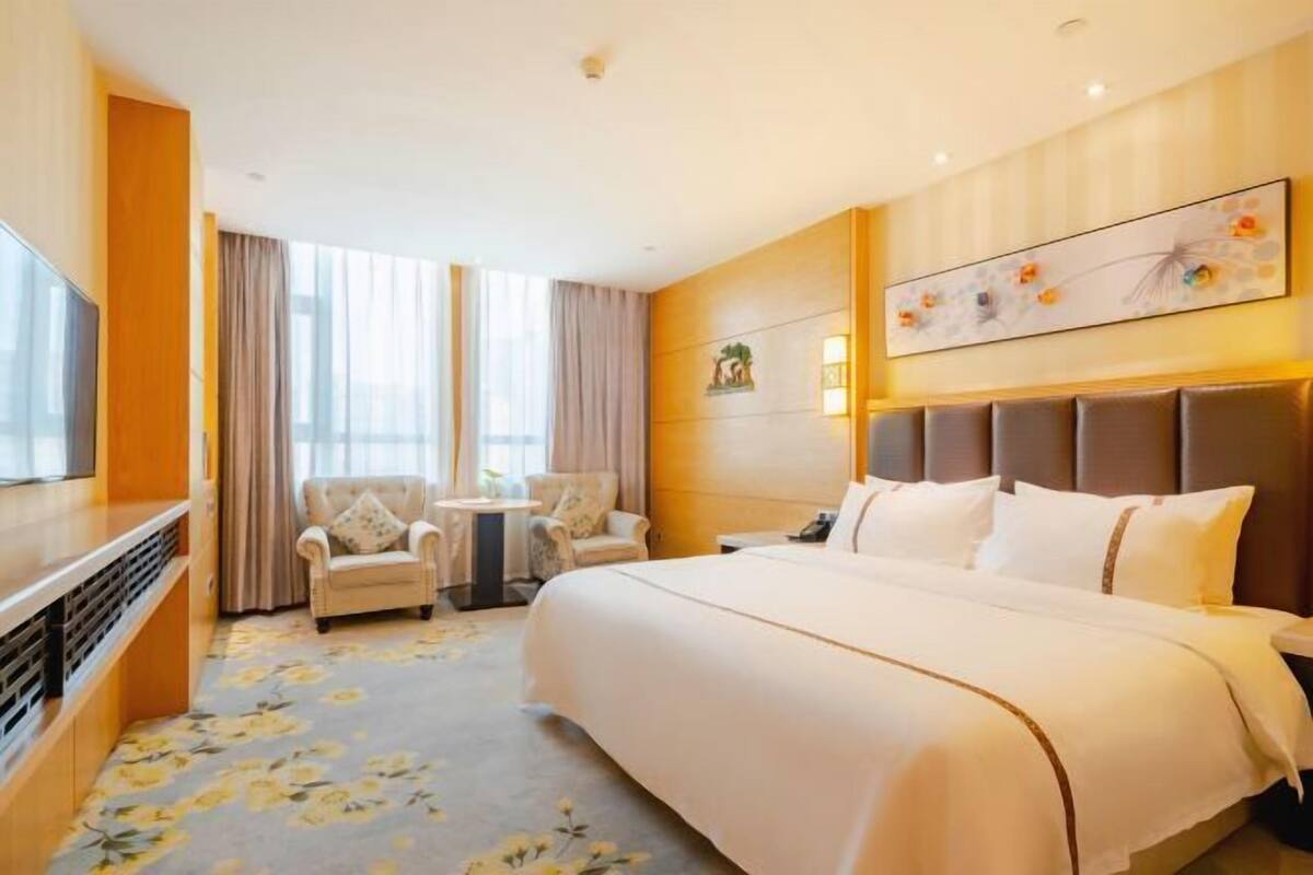 Quarto de hotel com uma cama grande, duas poltronas, uma mesinha, televisão e um quadro na parede.