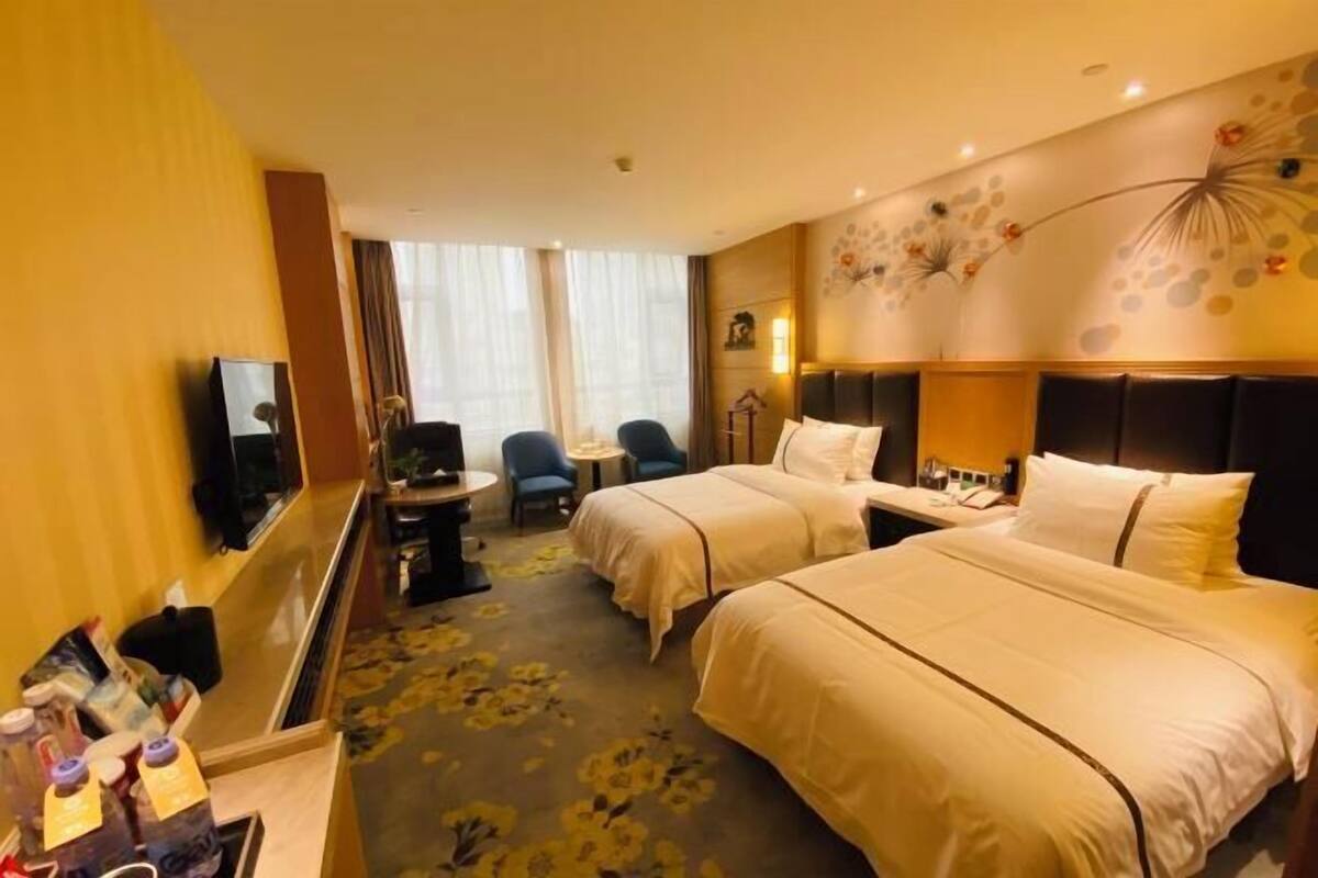 Quarto de hotel com duas camas, uma escrivaninha, uma cadeira, uma televisão e um detalhe decorativo na parede.