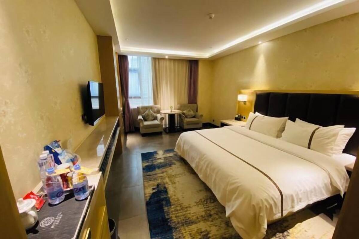 Quarto de hotel com uma cama grande, uma escrivaninha com luminária, televisão e área de estar com mesa e cadeiras.