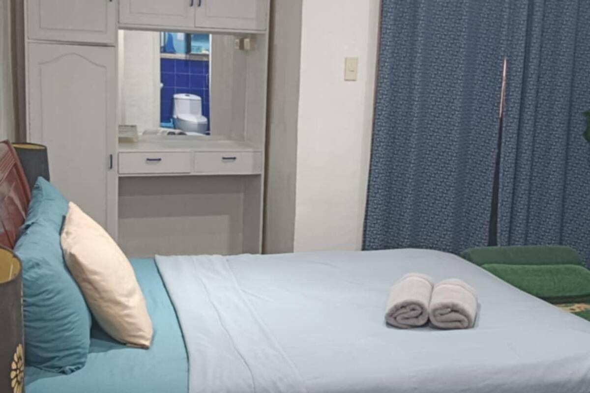Quarto com cama, mesa de cabeceira, espelho, armários e janela com cortinas.