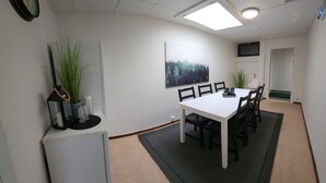Uma sala de conferências com uma mesa branca, cadeiras pretas, um relógio, uma planta e um quadro na parede.