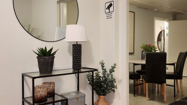 Um interior moderno com um espelho redondo, uma luminária, uma pequena planta e uma placa indicando uma câmera de segurança.