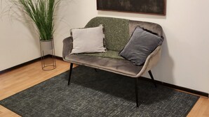 Uma sala de estar moderna com um sofá cinzento, um quadro abstrato grande e uma planta verde alta.