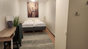 Quarto pequeno e bem iluminado, com uma cama, escrivaninha, abajur e uma janela grande.