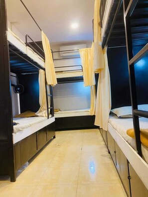 Um quarto estreito e bem iluminado, com beliches, uma mesa pequena e um ventilador.