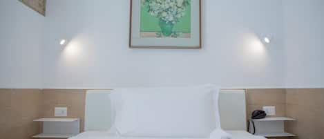 Quarto de hotel com cama, roupa de cama branca, pintura floral emoldurada na parede, mesas de cabeceira com abajures e um telefone em uma das mesas.