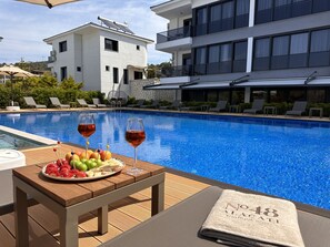 Uma área à beira da piscina com mesa de madeira posta com frutas e copos de vinho, cadeiras de descanso e uma toalha com a inscrição 'No48'.