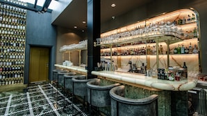 Uma área de bar com piso de mármore, diversas opções de assento e variedade de garrafas e copos nas prateleiras.