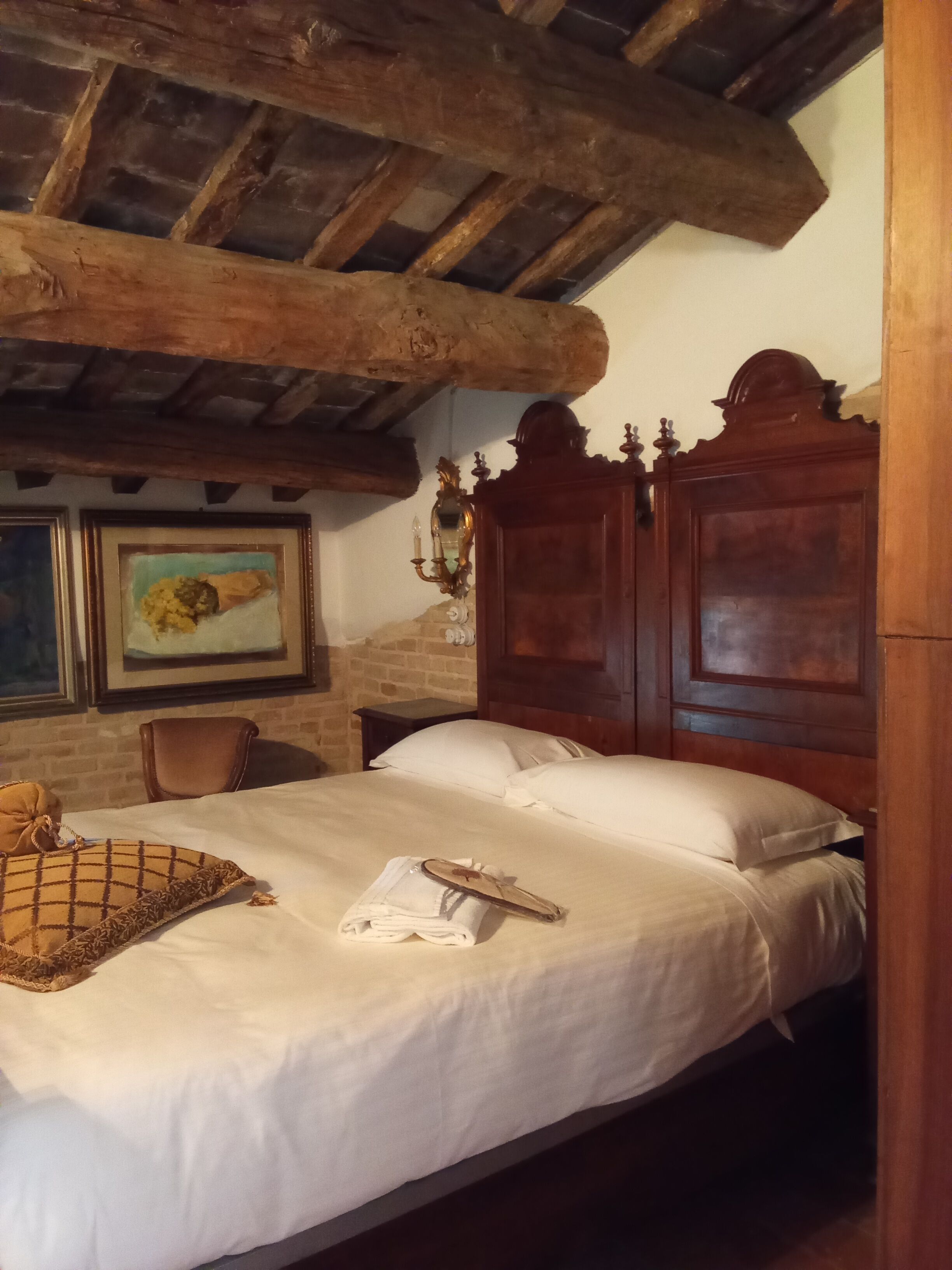 Quarto com cama de madeira, roupa de cama branca, cabeceira de madeira, pintura emoldurada e uma pequena mesa com abajur.