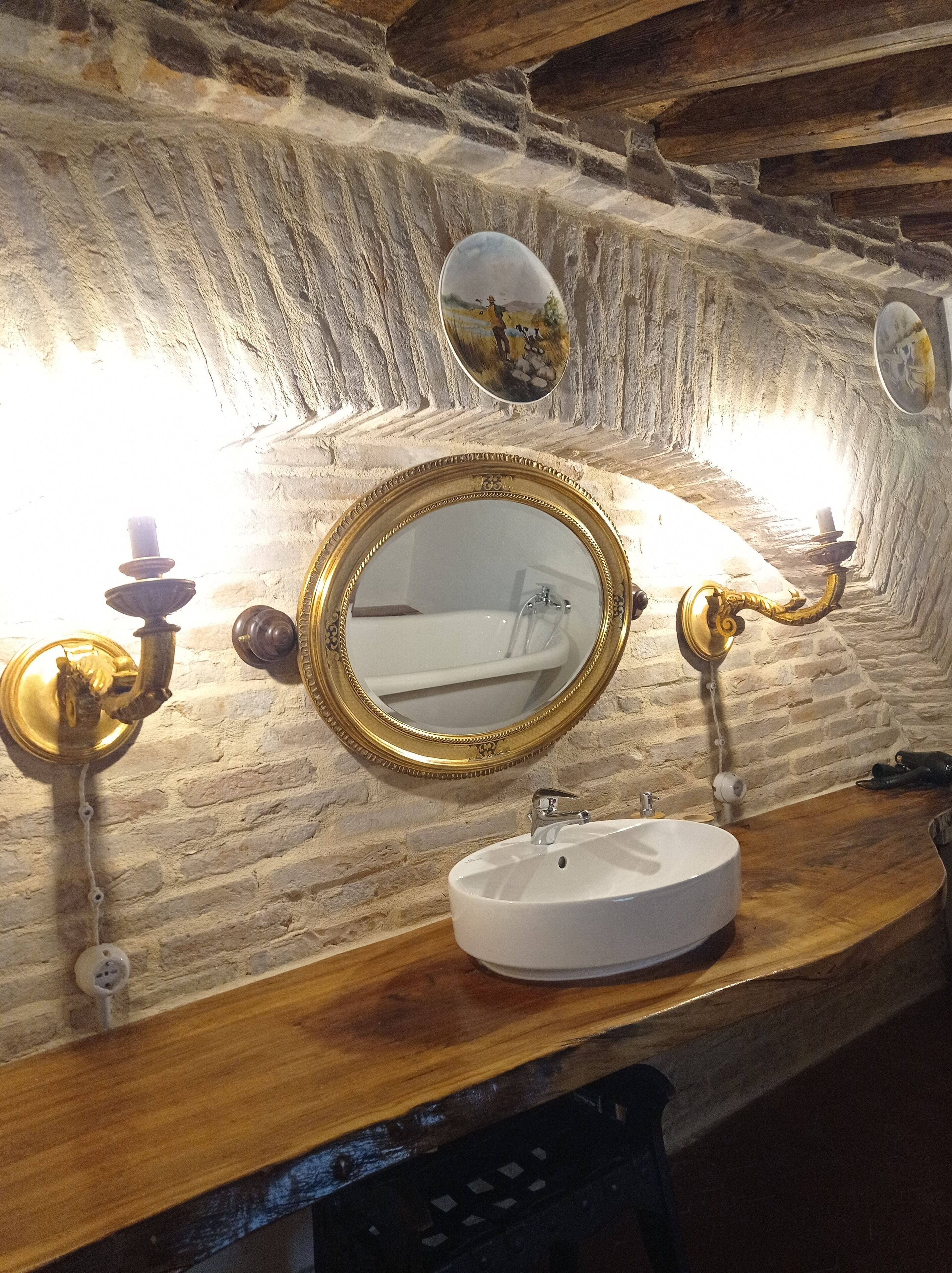 Casa de banho com parede de pedra, espelho redondo com moldura dourada, lavatório branco e duas candeeiros de parede com velas.