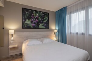 Quarto de hotel com uma cama grande, duas luminárias de cabeceira, uma obra de arte na parede e uma janela com cortinas.