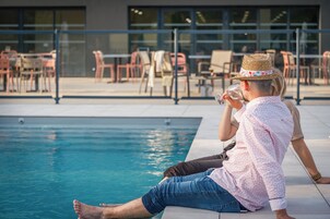 Uma área na beira da piscina com cadeiras e mesas, piscina e um edifício com grandes janelas ao fundo.