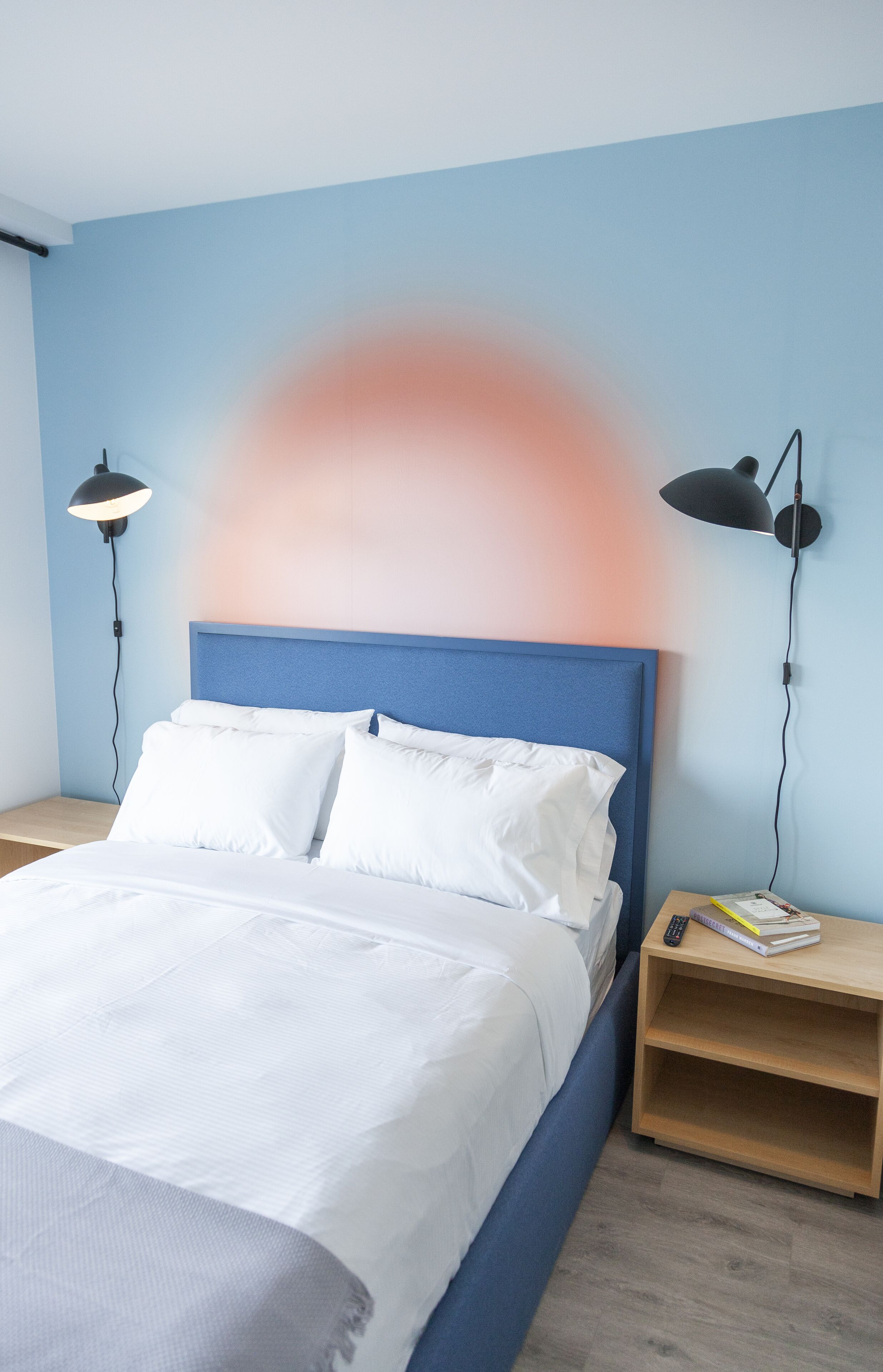 Cama bem arrumada com lençóis brancos, cabeceira azul e duas mesas de cabeceira com abajures.