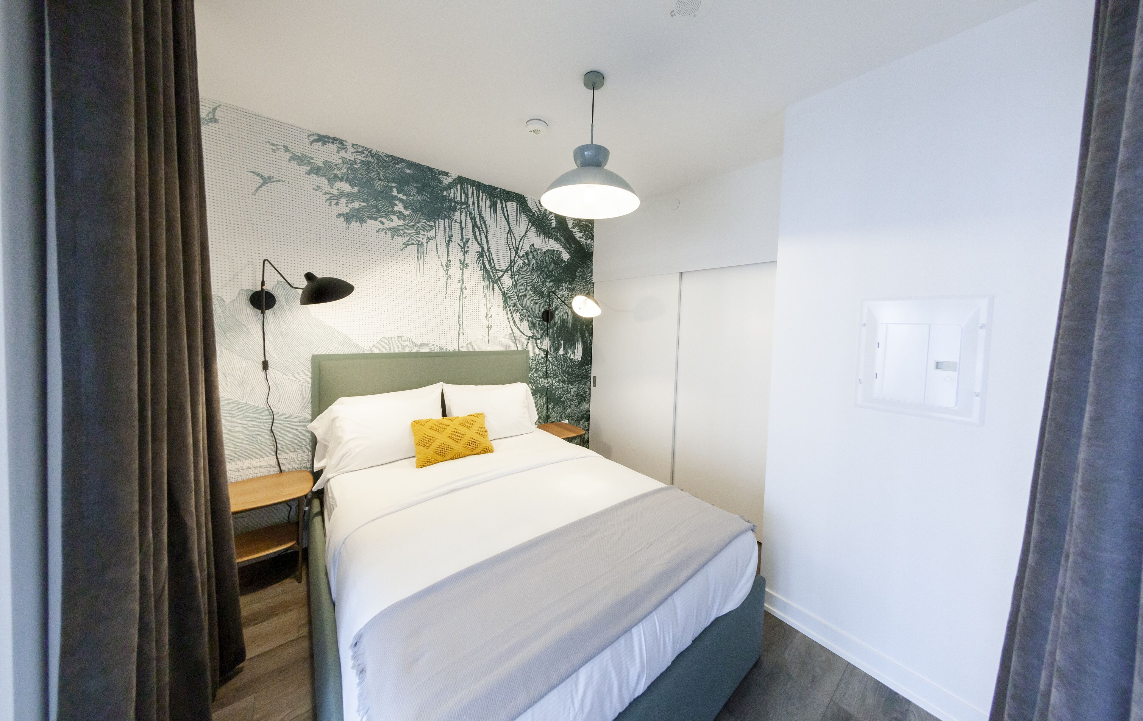 Quarto moderno com cama, mesa de cabeceira, luminária na parede e luz no teto.
