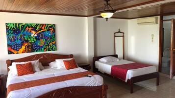 Quarto de hotel com duas camas de madeira, um quadro abstrato colorido na parede, um candeeiro no teto e uma porta de casa de banho visível.
