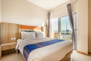 Quarto de hotel com uma cama grande, cabeceira de madeira, mesa de cabeceira e vista para o exterior através de uma janela ampla.