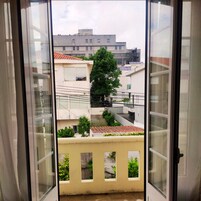 Vista de uma varanda com céu claro, uma árvore e edifícios ao fundo.