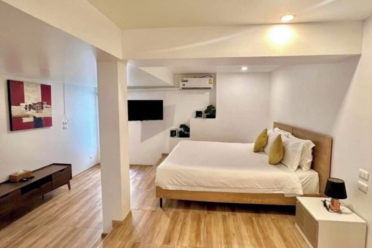 Quarto moderno com cama de madeira, roupa de cama branca, mesa de cabeceira com abajur e televisão fixada na parede.