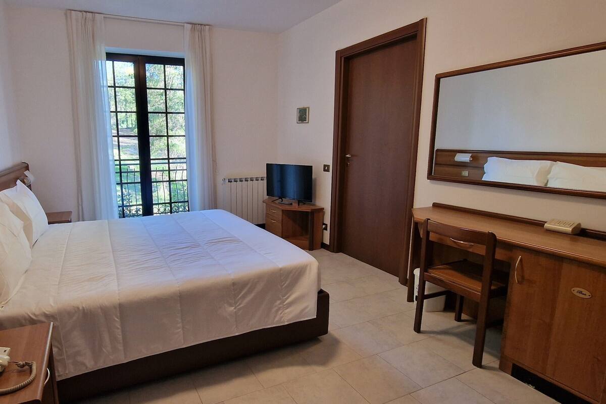 Quarto de hotel com cama, telefone ao lado da cama, escrivaninha com cadeira, televisão e janela com cortinas.