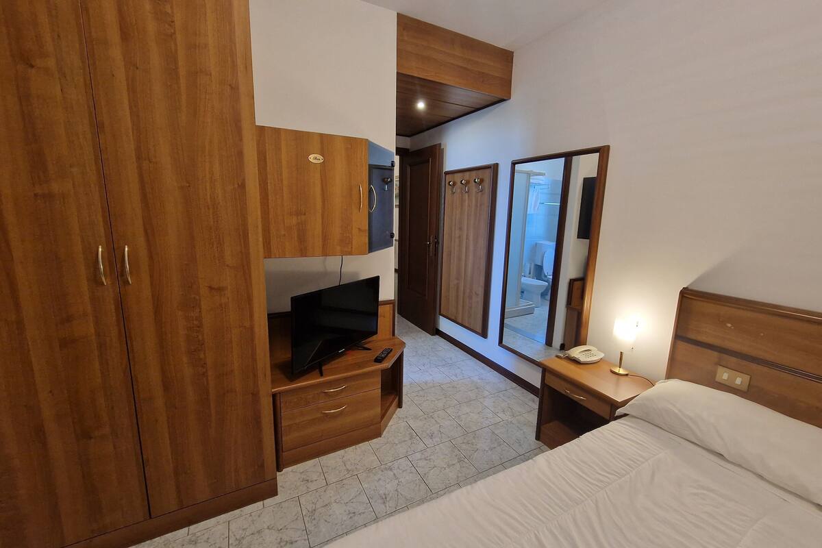 Quarto com armário de madeira, televisão sobre suporte de madeira, mesa de cabeceira com abajur e um espelho grande.