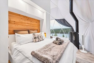 Quarto moderno com cabeceira de madeira, cama grande com roupa de cama branca, manta macia e vista para um lago através de uma estrutura de janela geométrica.