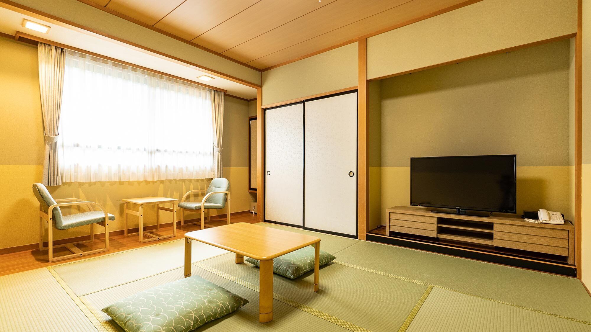 Um quarto japonês tradicional com tatames, uma mesa baixa, duas cadeiras, uma televisão e uma porta deslizante.