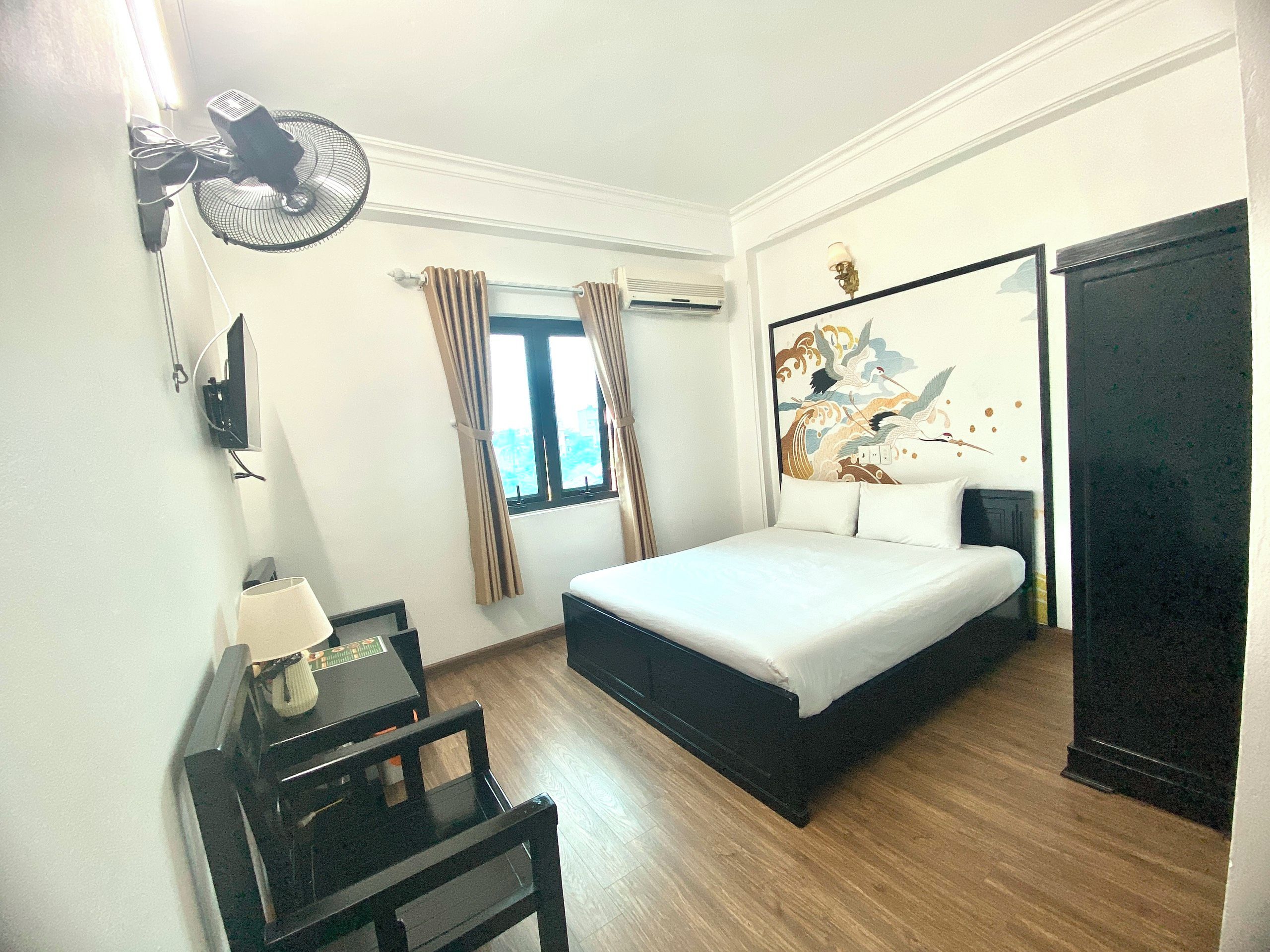 Quarto de hotel com cama, escrivaninha, cadeira, ventoinha, janela com cortinas e ar condicionado fixado na parede.