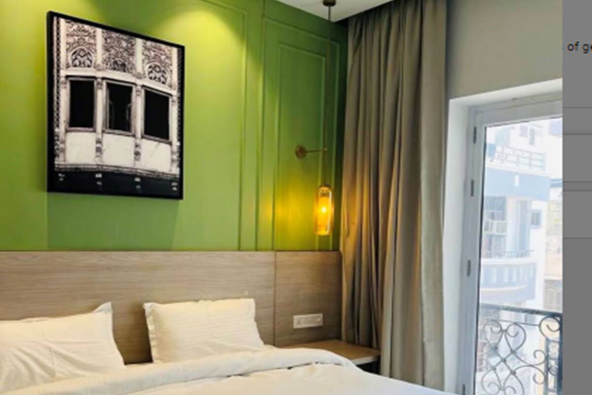 Quarto de hotel com cama, parede verde, quadro emoldurado, mesa de cabeceira, abajur e janela com cortinas.